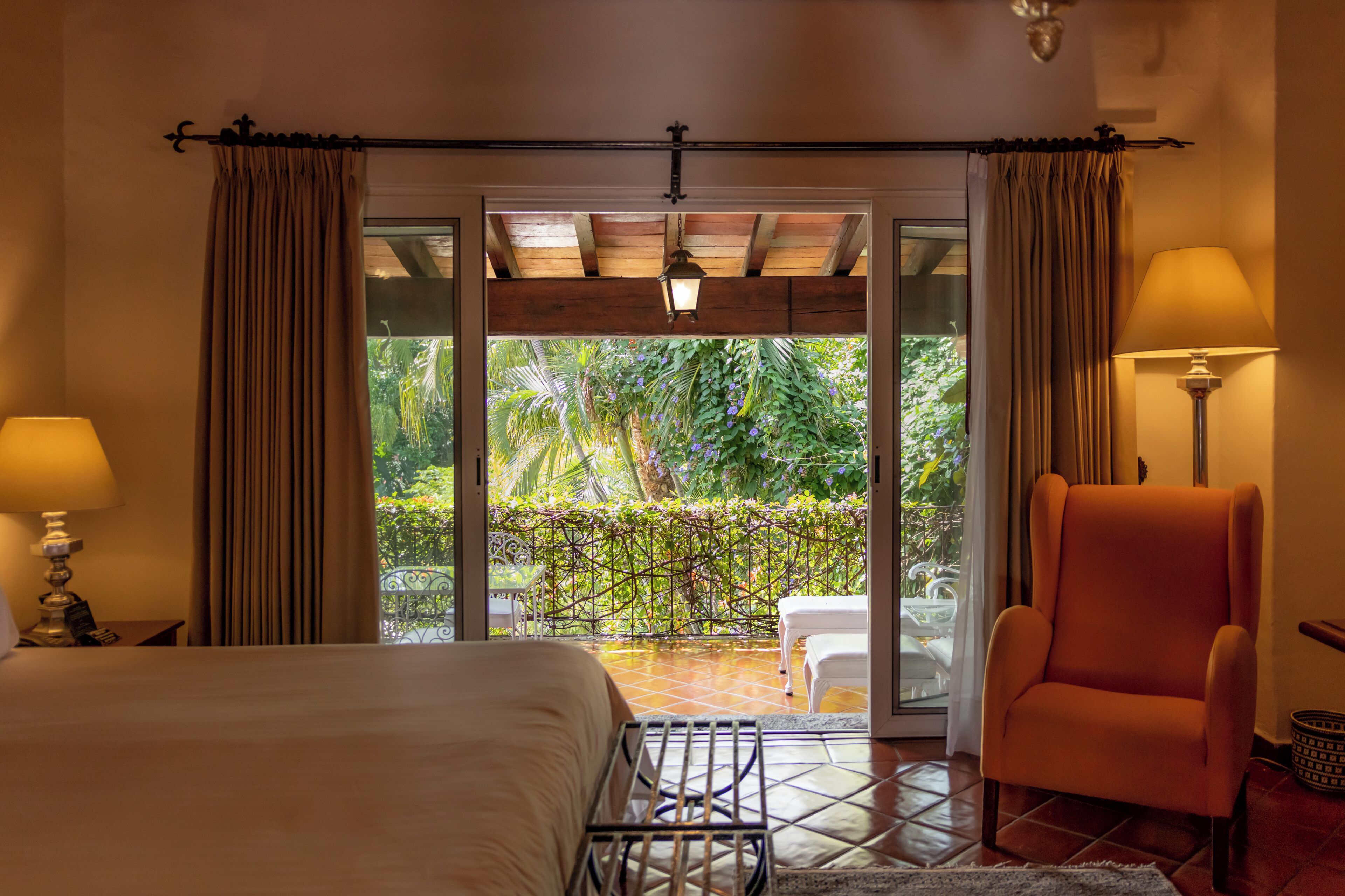 Quarto de hotel com cama, cadeira, mesa e vista para um jardim através de uma porta de vidro.