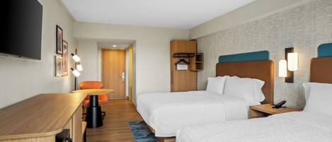 Quarto de hotel com duas camas, uma escrivaninha, uma cadeira, uma televisão e uma prateleira na parede.