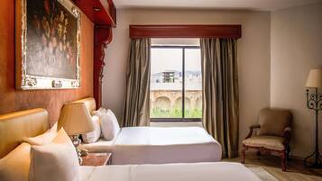 Quarto de hotel com duas camas, um quadro na parede, vista de um edifício pela janela e uma cadeira.
