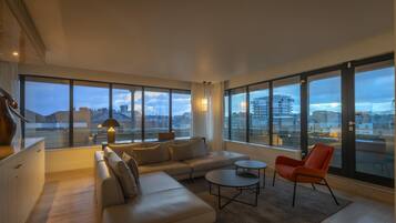 Uma sala de estar moderna com sofá, poltrona e mesa de centro, janelas amplas com vista para a cidade e um abajur.
