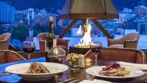 Uma mesa de jantar posta com pratos de comida, copos de vinho e uma garrafa de vinho. Um braseiro é visível ao fundo.