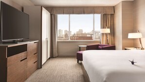 Quarto de hotel com uma cama, televisão, escrivaninha e vista para o horizonte da cidade.