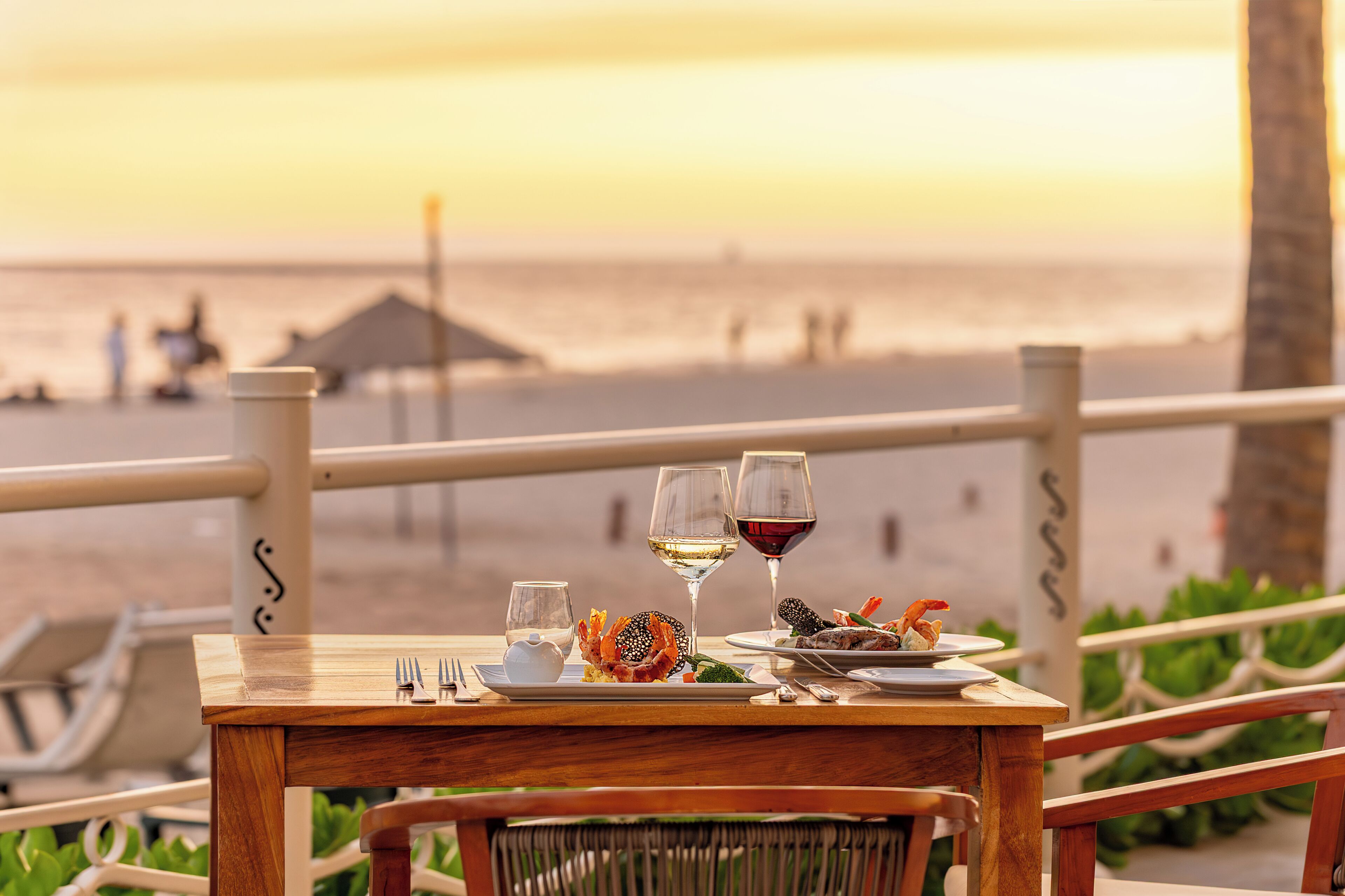 Uma mesa de madeira com pratos de comida e copos de vinho, com vista para uma praia onde se veem pessoas ao longe.