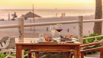 Uma mesa de madeira com pratos de comida e copos de vinho, com vista para uma praia onde se veem pessoas ao longe.