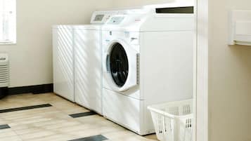 Uma lavanderia com duas máquinas de lavar frontais empilhadas, uma cesta de roupa branca e um ventilador à esquerda.