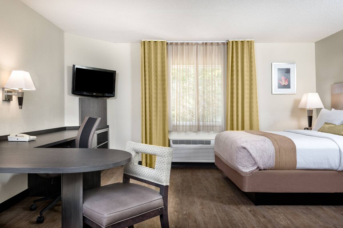 Quarto de hotel com cama, escrivaninha, cadeira, televisão e janela com cortinas.
