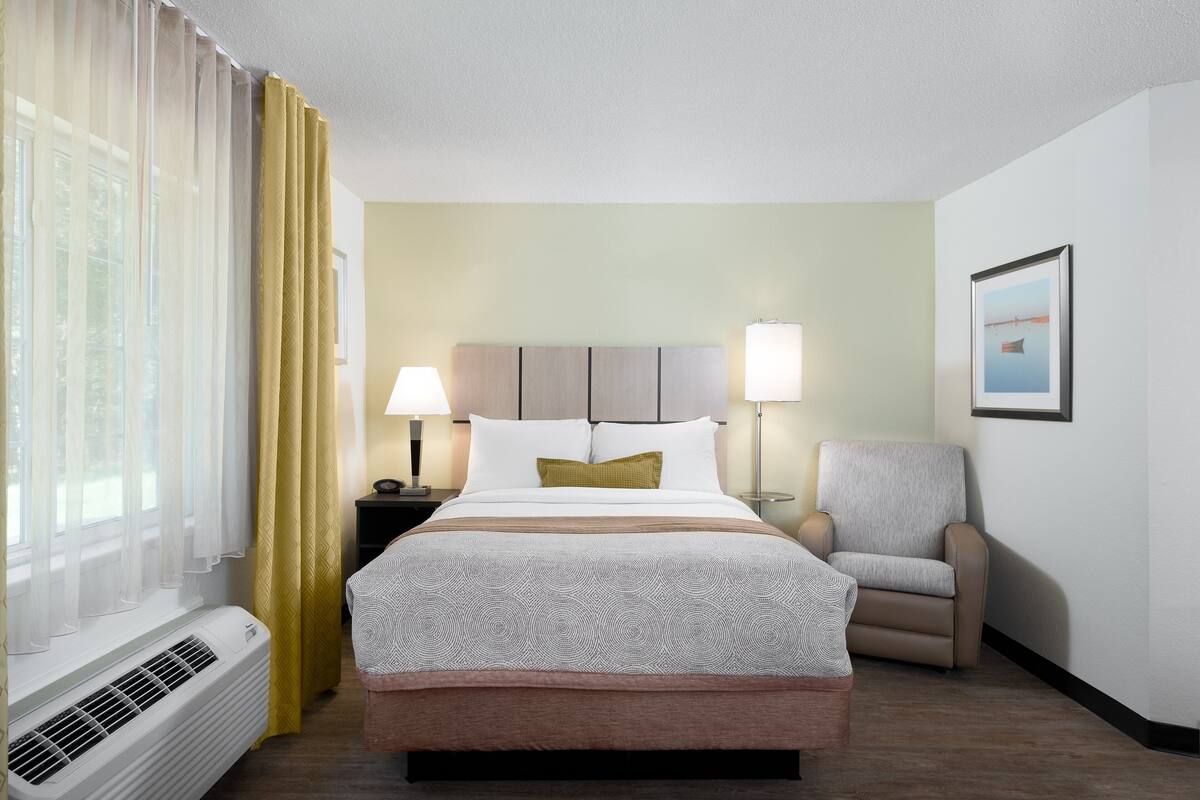 Quarto de hotel com cama, duas luminárias de cabeceira, uma poltrona, janela com cortinas e um quadro na parede.