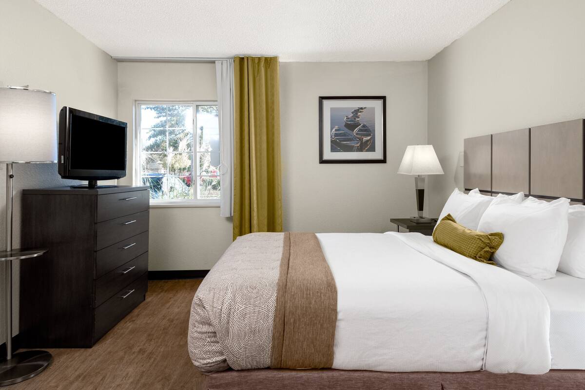 Quarto de hotel com uma cama, televisão, escrivaninha, luminária e janela com cortinas.