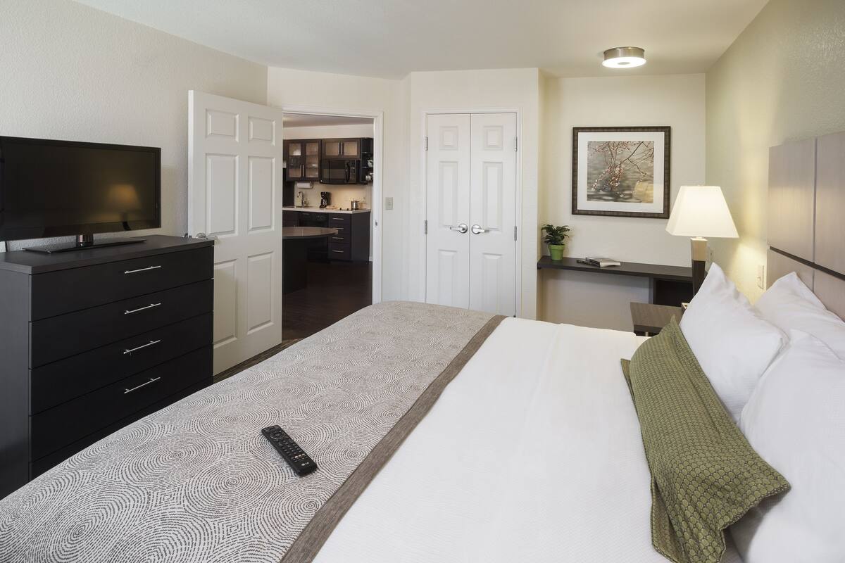 Quarto de hotel com cama, penteadeira, televisão, escrivaninha com abajur e vista para a cozinha.