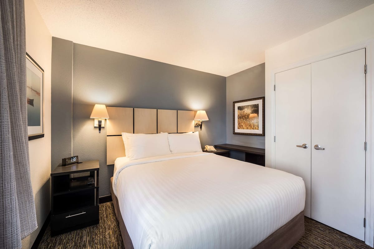 Quarto de hotel com uma cama grande, duas mesas de cabeceira com abajures, um criado-mudo, um guarda-roupa e um quadro emoldurado na parede.