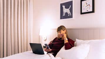 Uma pessoa sentada em uma cama, com um laptop e um smartphone, uma moldura com a foto de um cão e uma obra de arte abstrata emoldurada na parede.