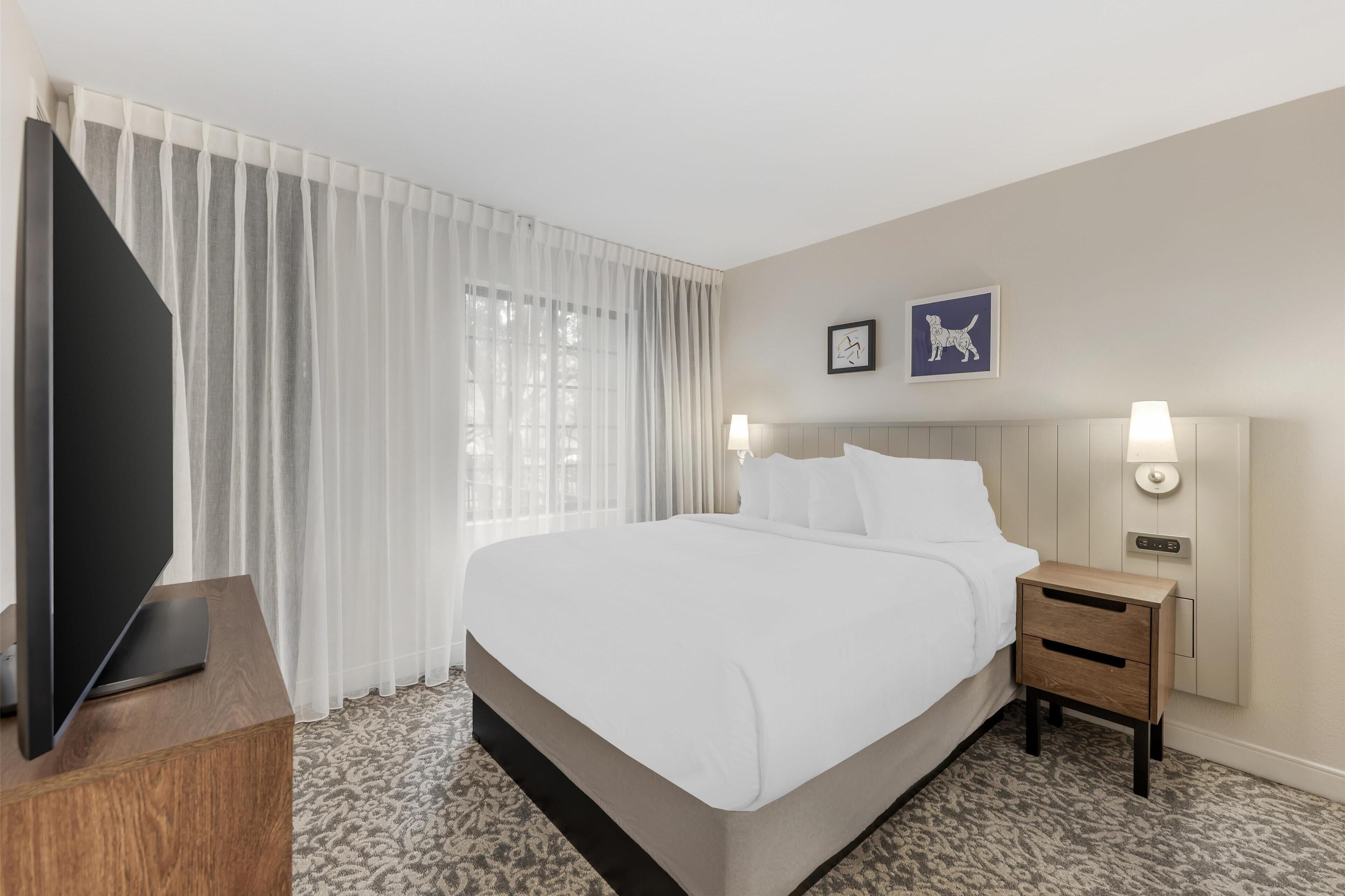 Quarto de hotel com uma cama grande, uma televisão em suporte de madeira, mesa de cabeceira com abajur e janela com cortinas leves.