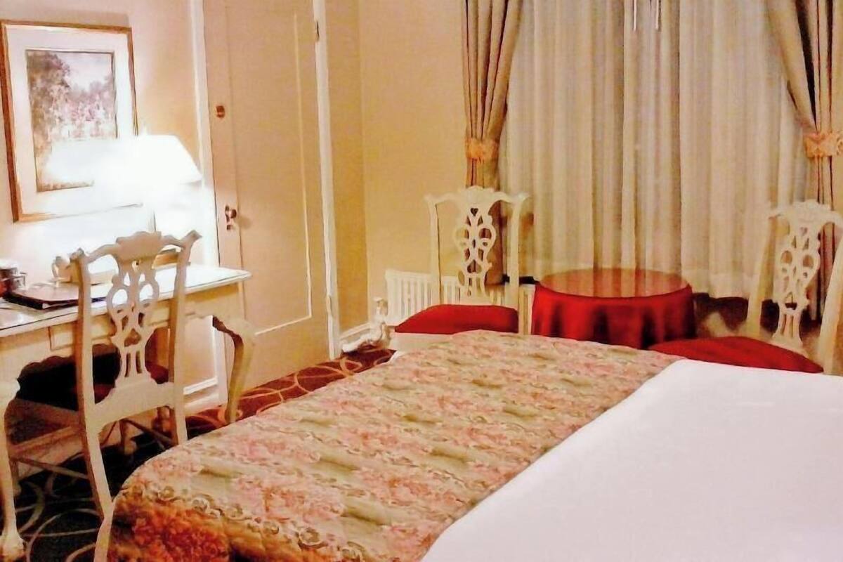 Quarto de hotel com cama, escrivaninha, duas cadeiras, uma mesinha e cortinas.