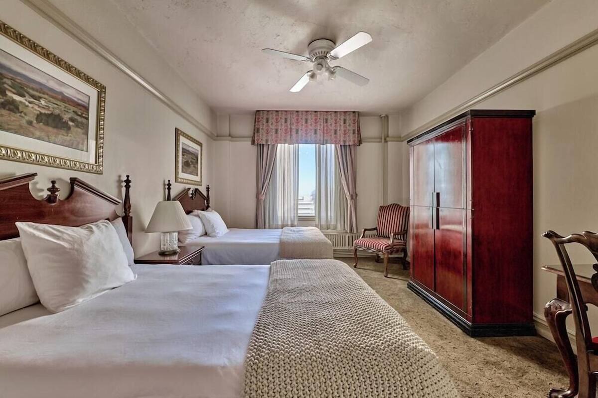 Quarto de hotel com duas camas, um ventilador de teto, um guarda-roupa vermelho grande e uma janela com cortinas.