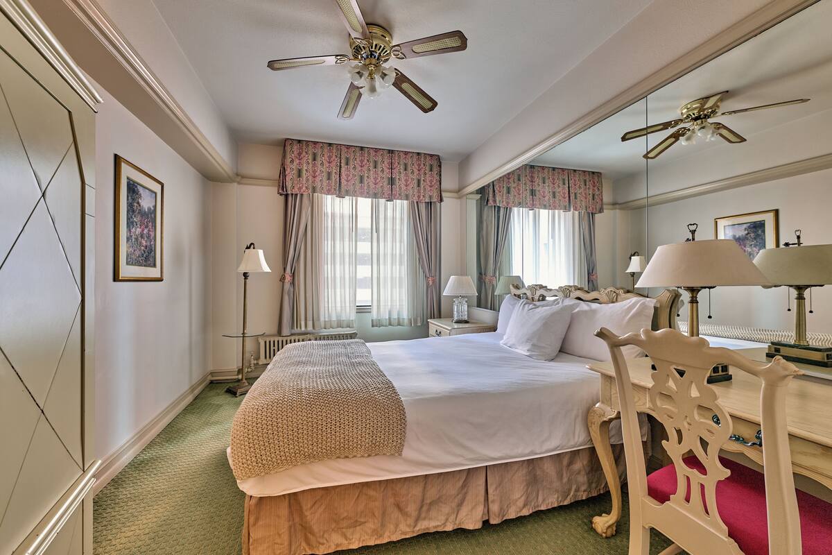 Quarto de hotel com uma cama grande, um ventilador de teto, uma escrivaninha, uma cadeira e um espelho.