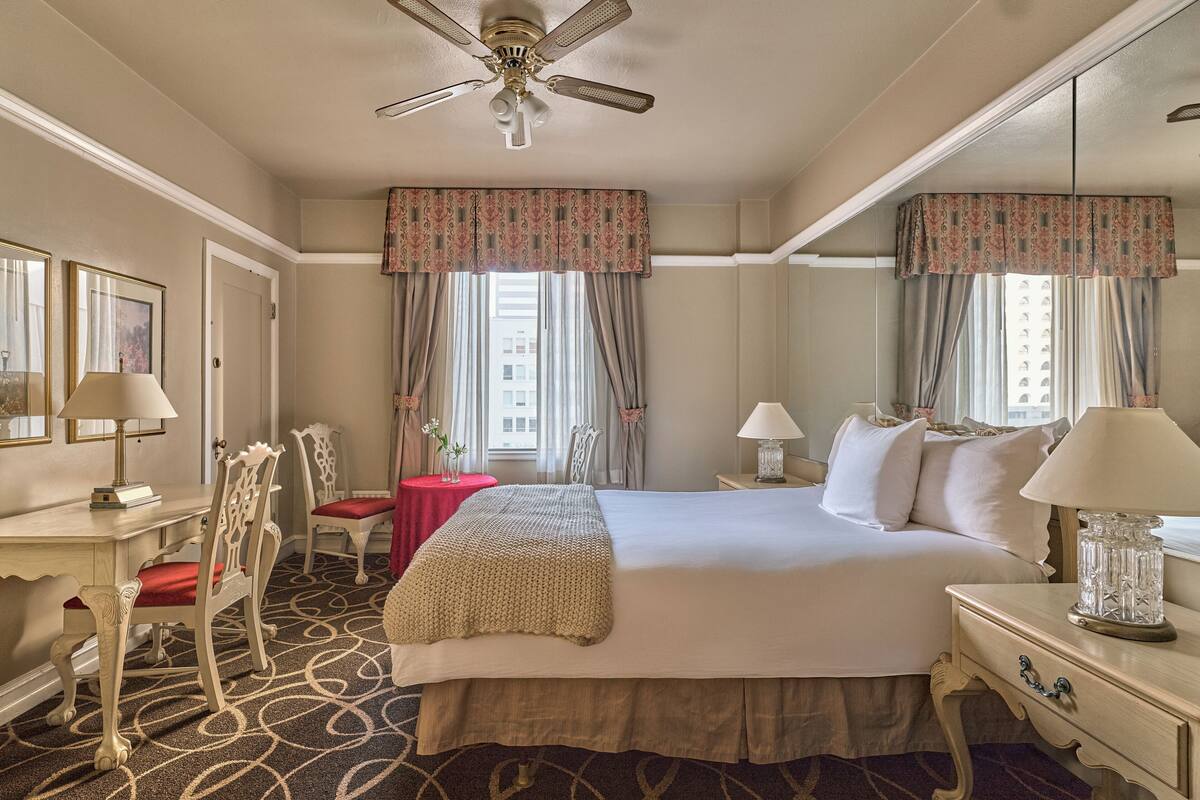 Quarto de hotel com uma cama grande, um ventilador de teto, uma escrivaninha com abajur e uma cadeira.