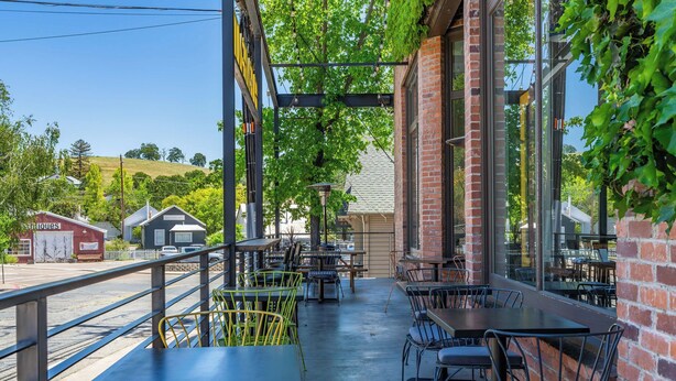 Um café ao ar livre com mesas e cadeiras, um muro de tijolos e vista para a rua.