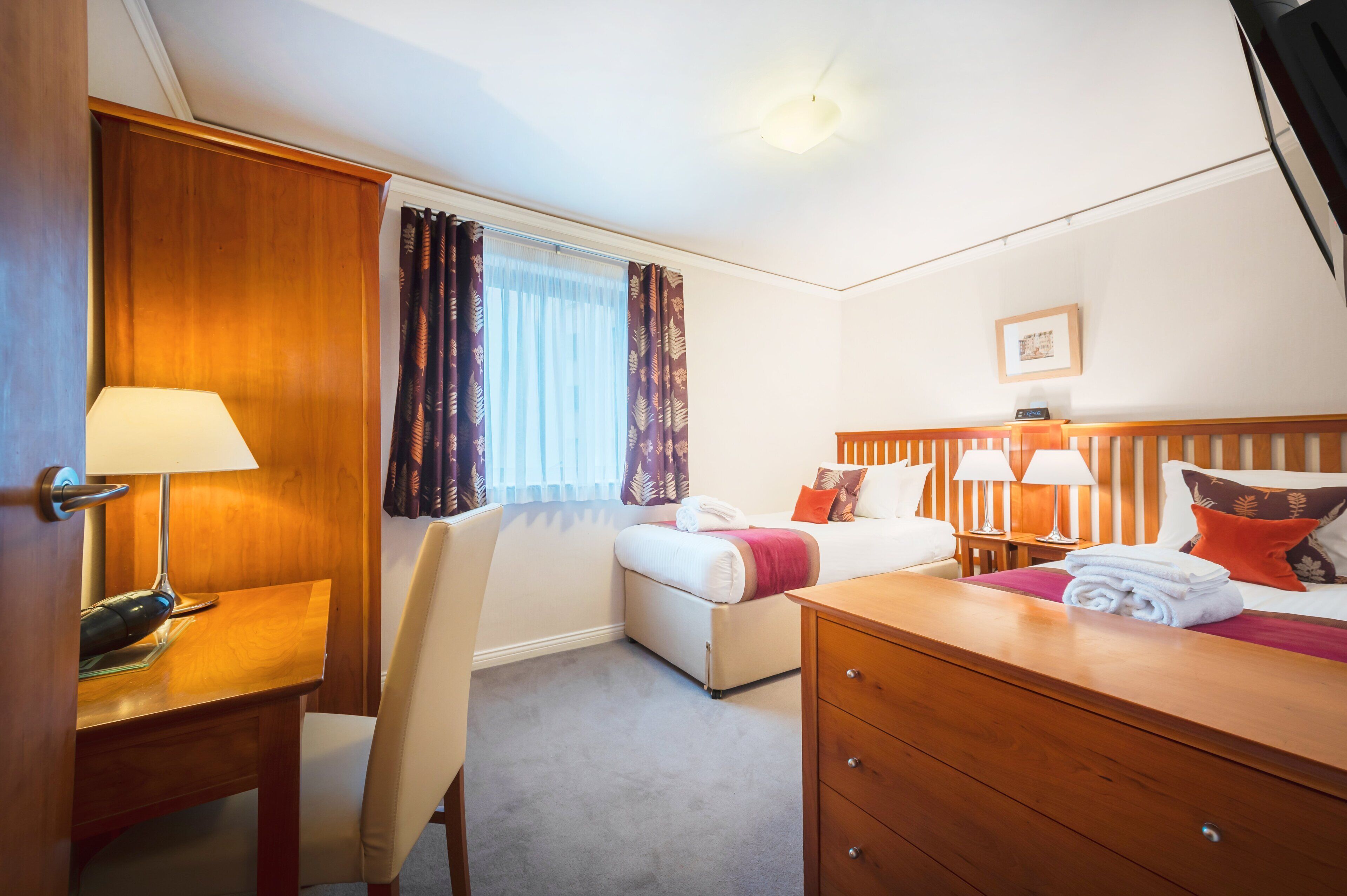 Quarto de hotel com cama, mesas de cabeceira, escrivaninha, cadeira, guarda-roupa e janela com cortinas.
