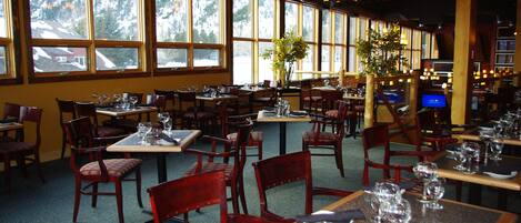 Um restaurante com várias mesas e cadeiras, grandes janelas com vista para montanhas cobertas de neve e luminárias pendentes em estilo de lanterna.