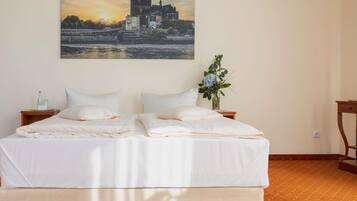 Uma cama de casal com roupa de cama branca, uma mesa de cabeceira com uma garrafa e um copo, um vaso com flores e um quadro de um castelo na parede.