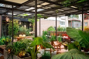 Um alpendre coberto com móveis de madeira, plantas em vasos e um muro de tijolos ao fundo.