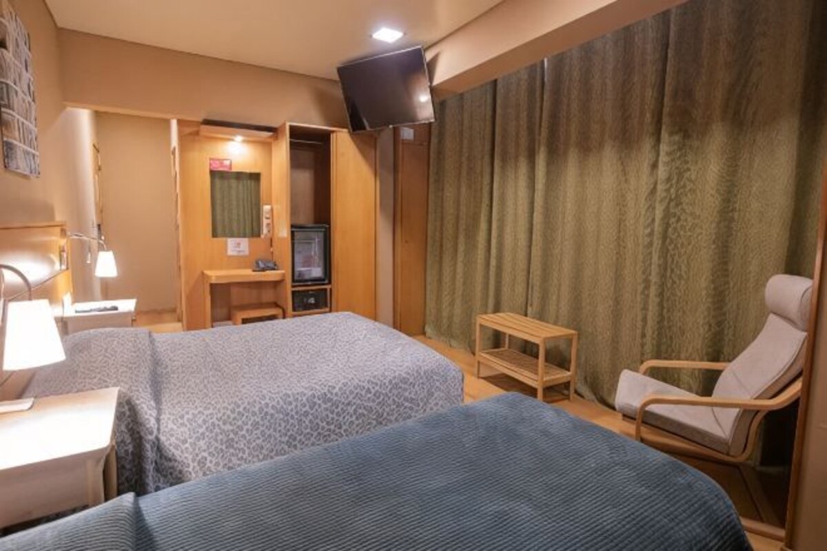 Quarto de hotel com duas camas, televisão, escrivaninha, cadeira e cortina.
