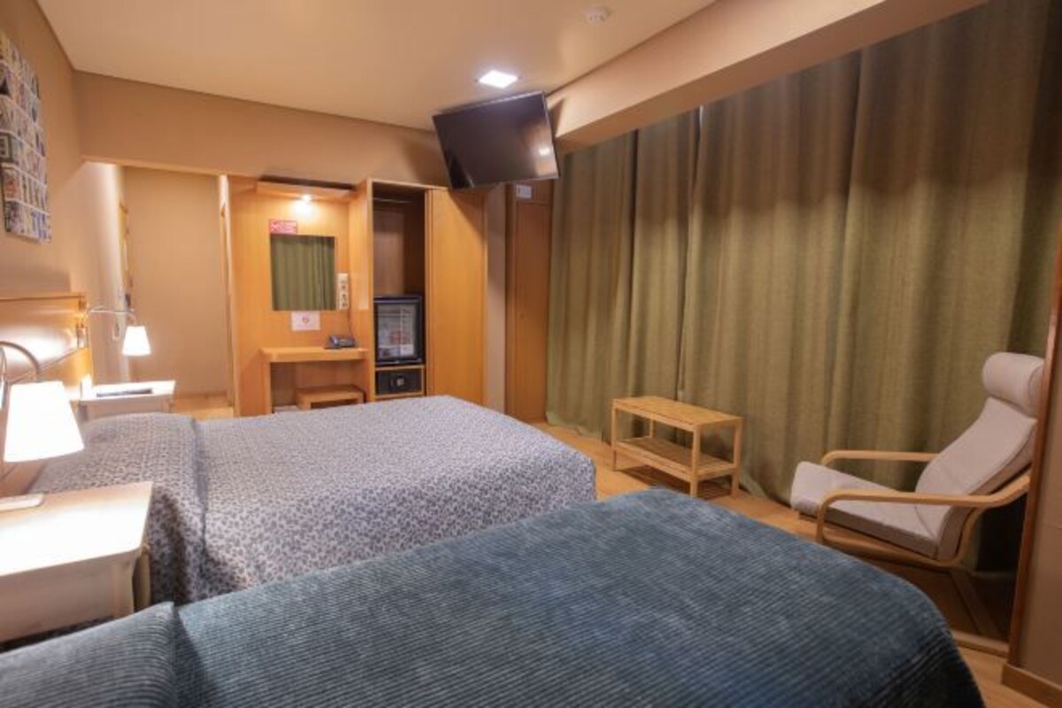 Quarto de hotel com duas camas, uma escrivaninha, uma cadeira, uma televisão e uma cortina.