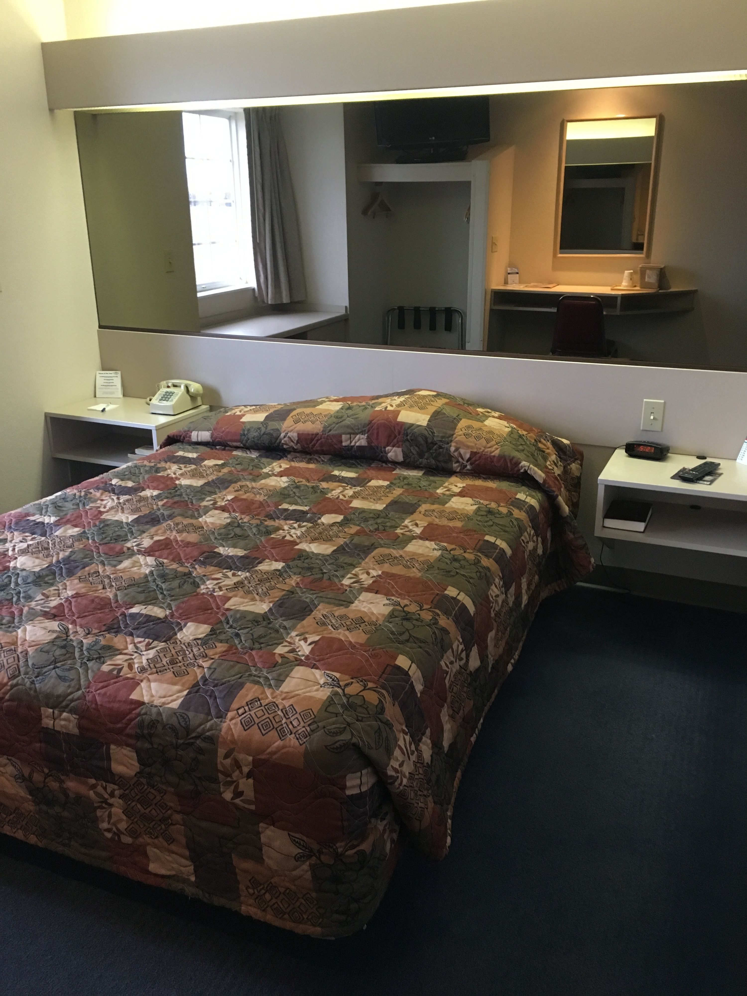 Quarto de hotel com cama grande, escrivaninha com telefone, espelho e mesinha de apoio com relógio e controle remoto.