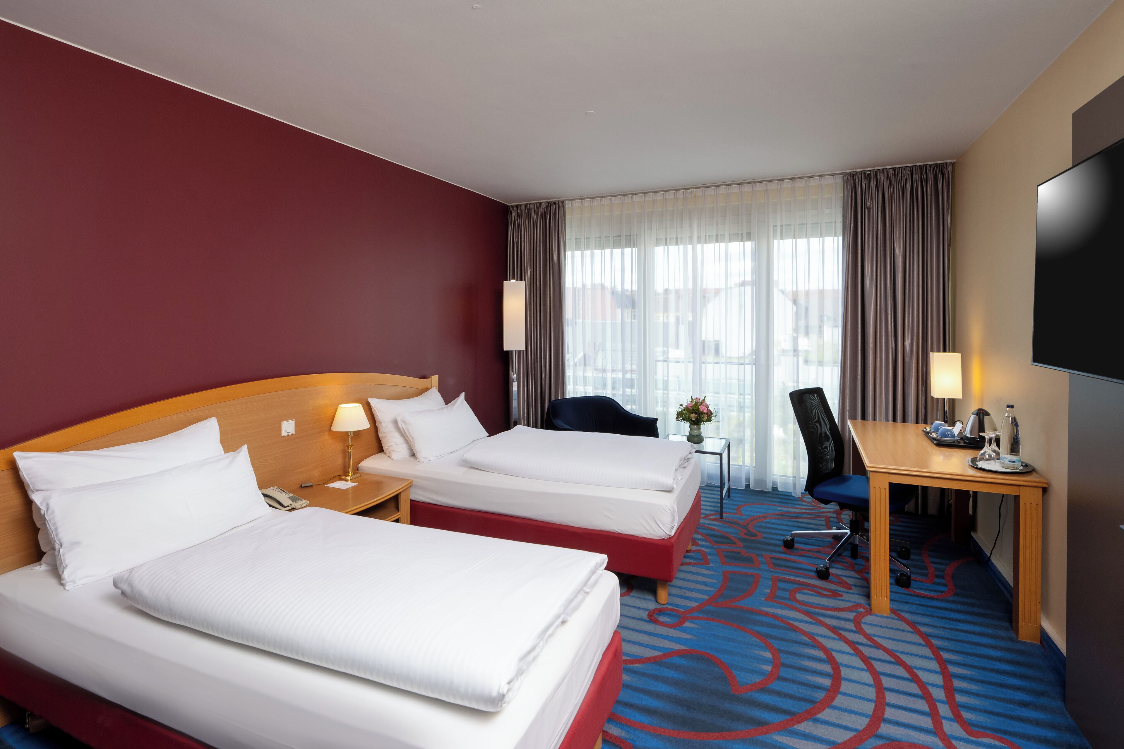 Quarto de hotel com duas camas, uma escrivaninha, uma cadeira, televisão e janela com cortinas.