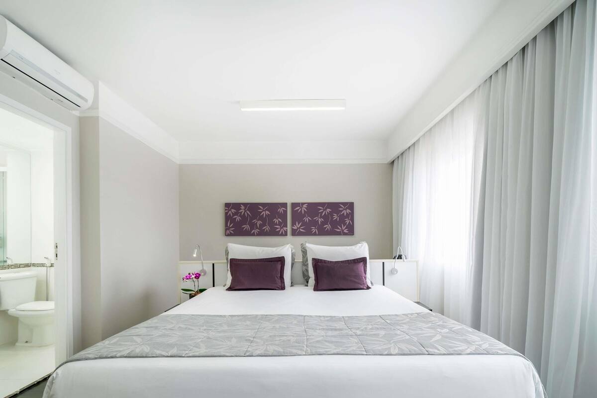 Quarto de hotel com uma cama grande, mesa de cabeceira com luminária, espelho e janela com cortinas.