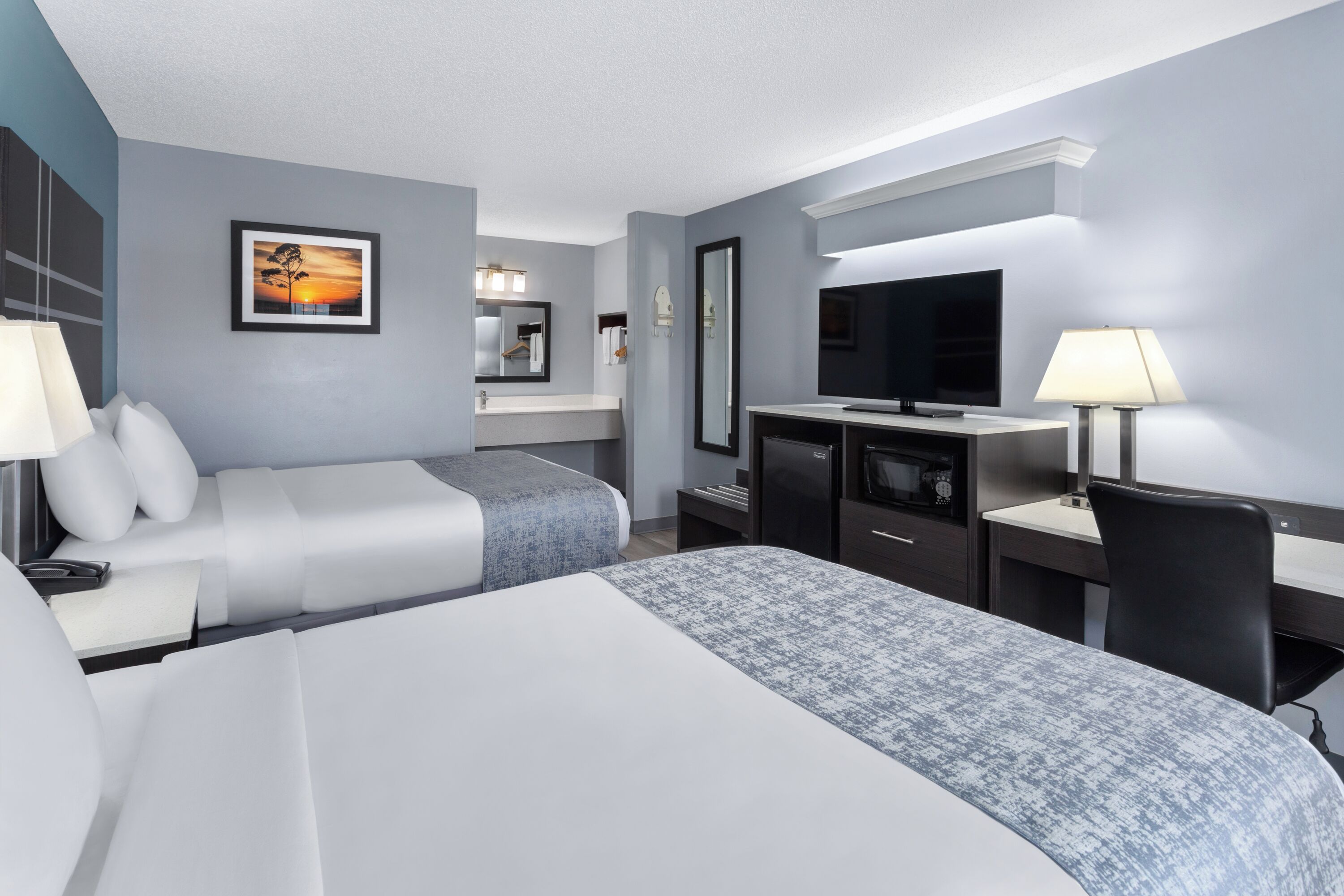 Quarto de hotel com duas camas, uma escrivaninha, uma televisão e banheiro com espelho e porta-toalhas.