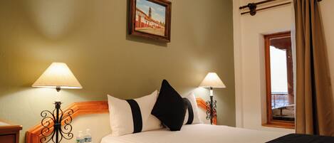 Quarto de hotel com cama, duas luminárias de cabeceira, mesa de apoio, quadro na parede e janela com cortinas.