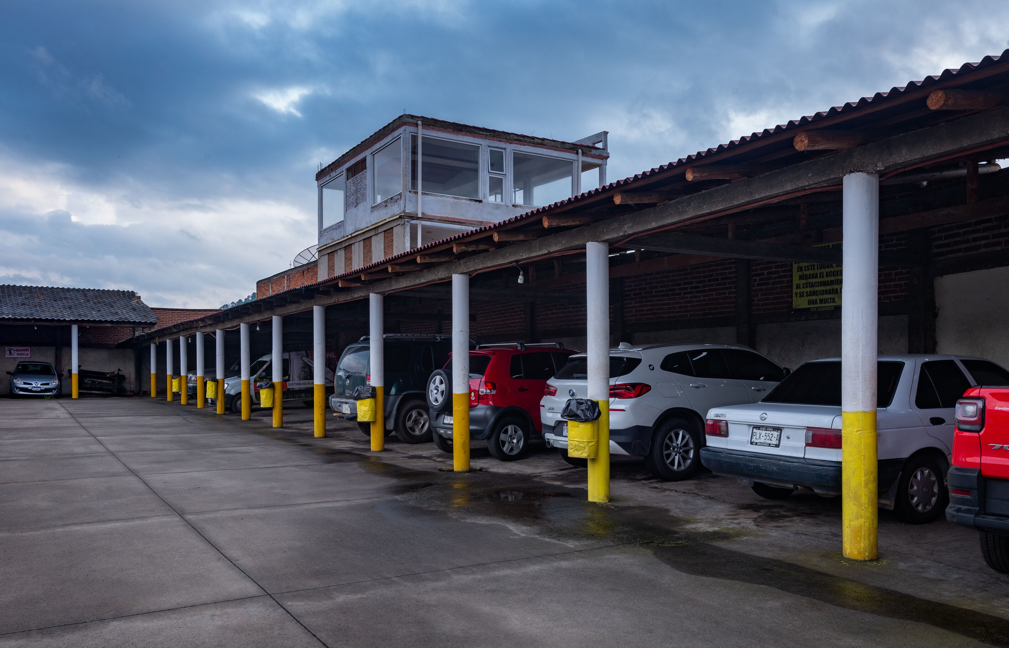 Uma garagem de estacionamento com vários carros estacionados sob uma área coberta, com parede de tijolos e teto de metal.