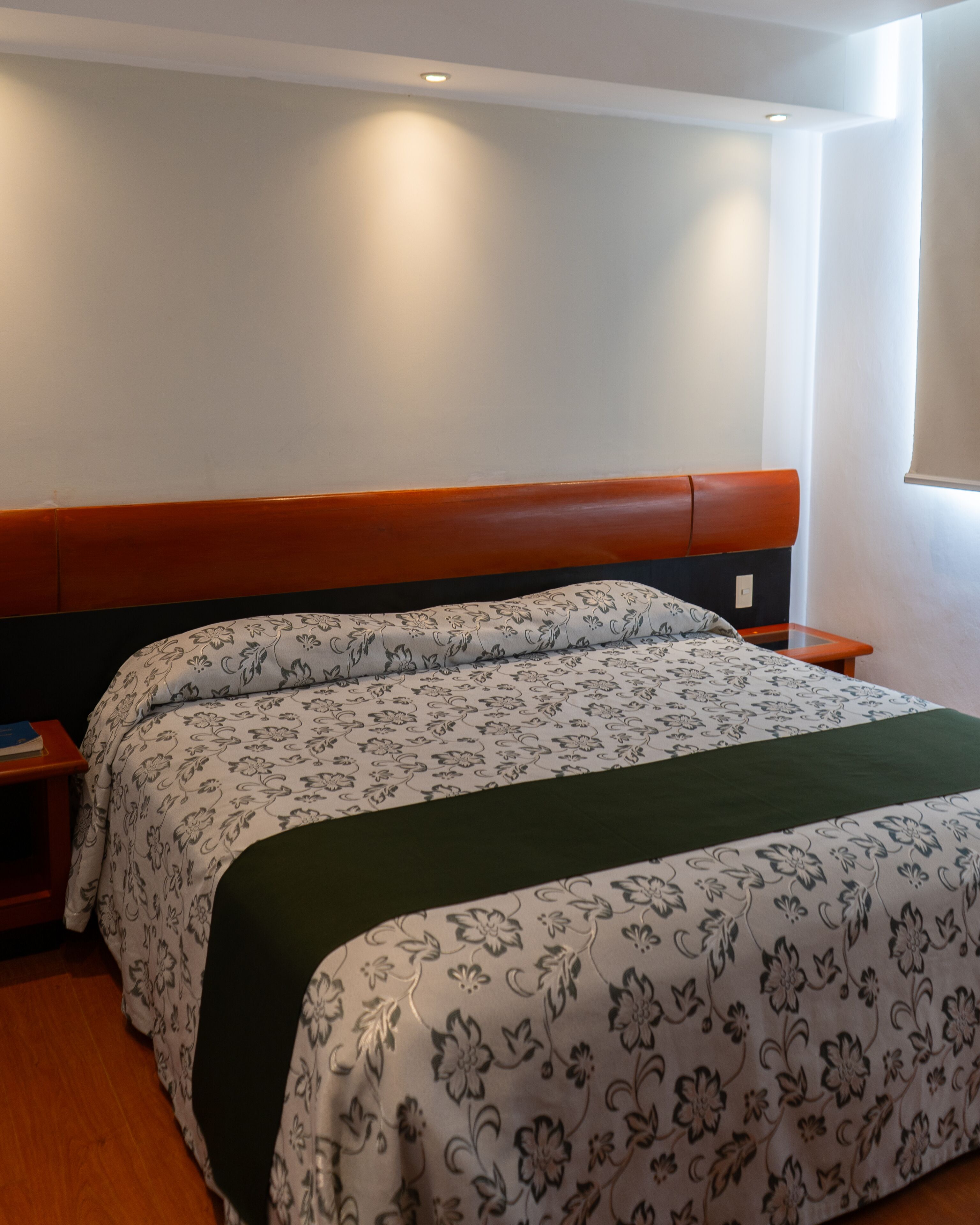 Quarto com cabeceira de madeira, roupa de cama florida e mesa de cabeceira com um livro.
