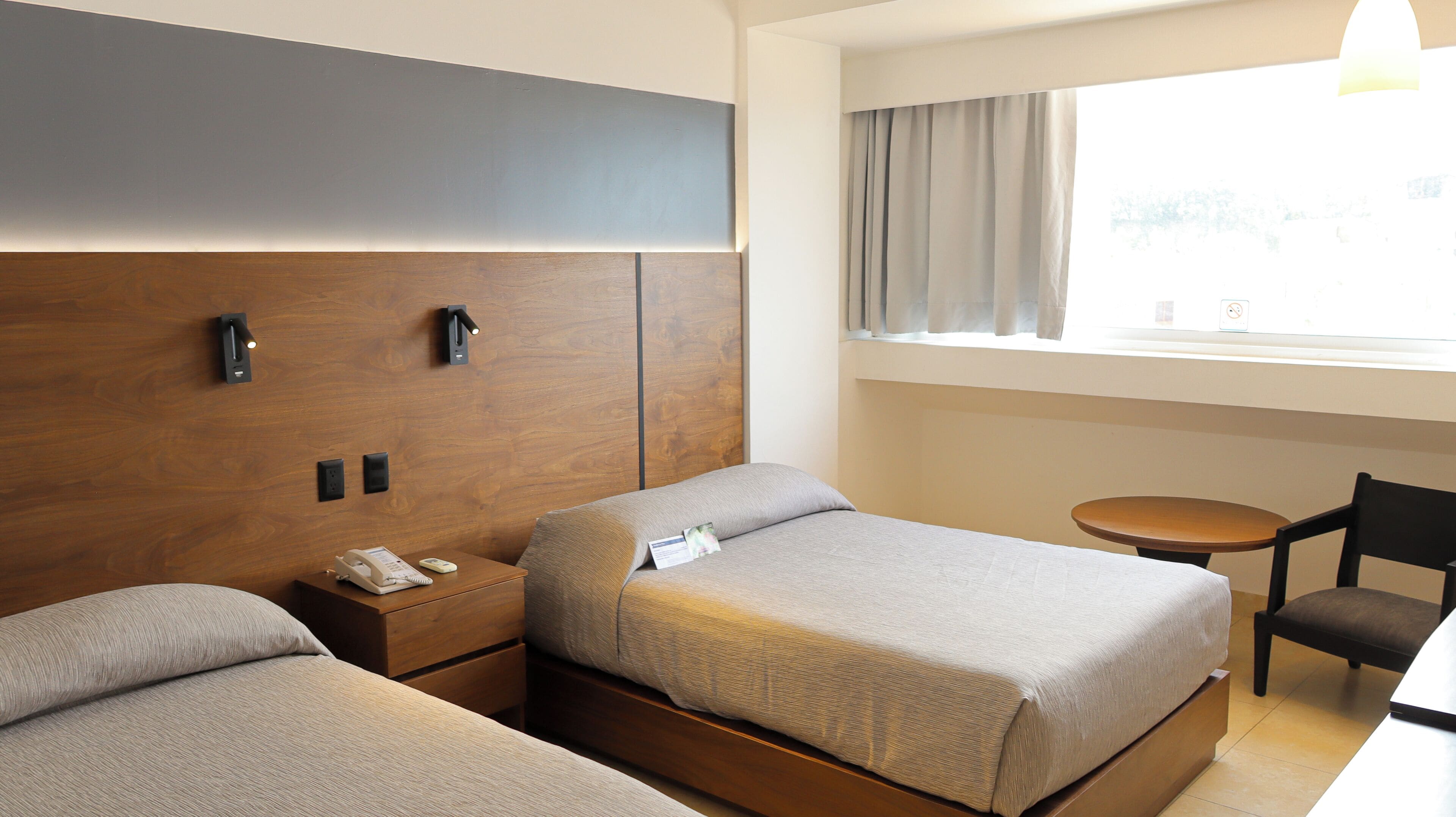 Quarto de hotel com duas camas, cabeceira de madeira, mesas de cabeceira, escrivaninha, cadeira e janela com cortinas.