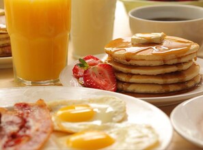 Uma mesa de café da manhã com panquecas, ovos, bacon e um copo de suco de laranja.