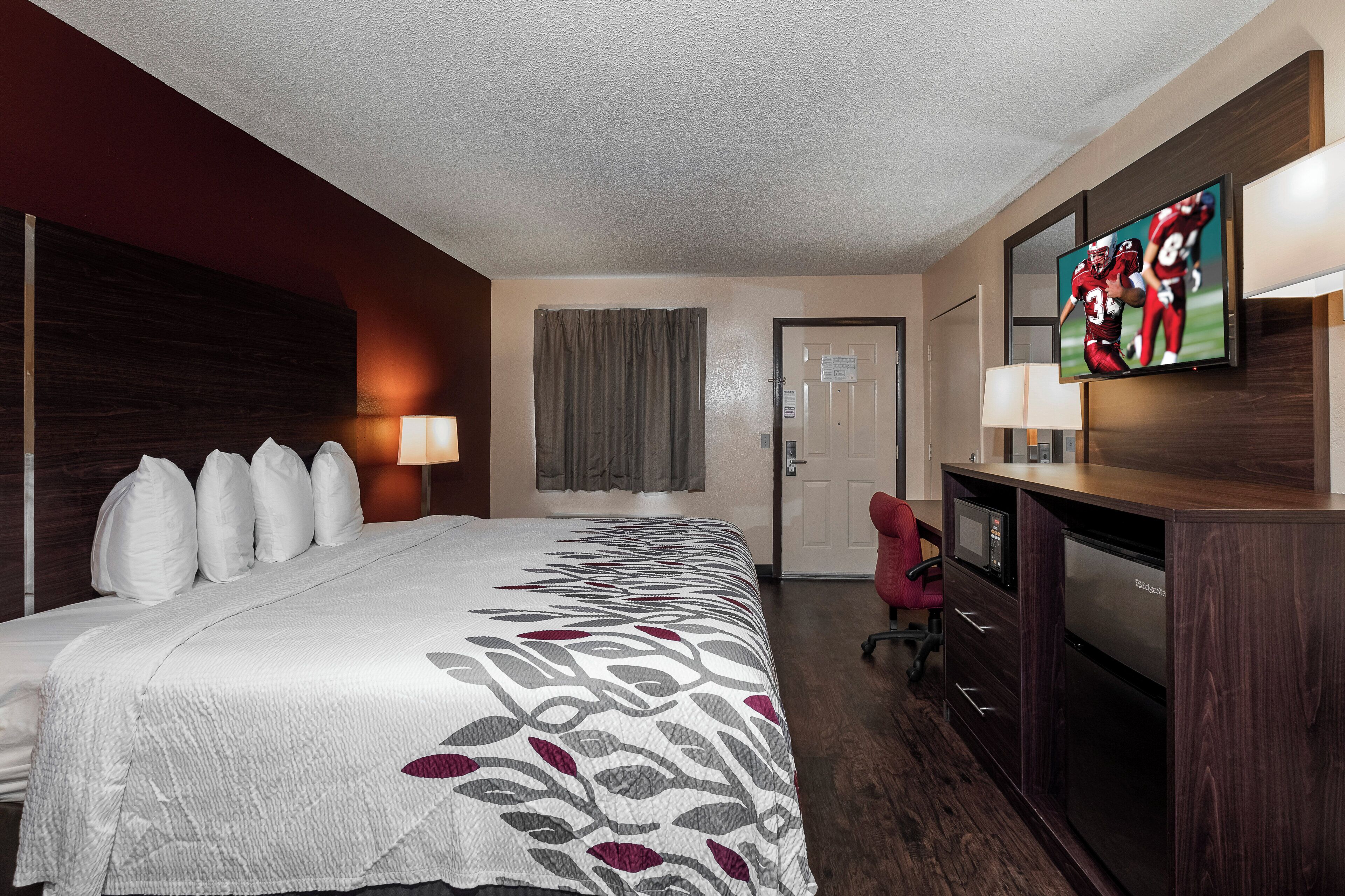 Quarto de hotel com cama, escrivaninha com televisão, micro-ondas e porta.