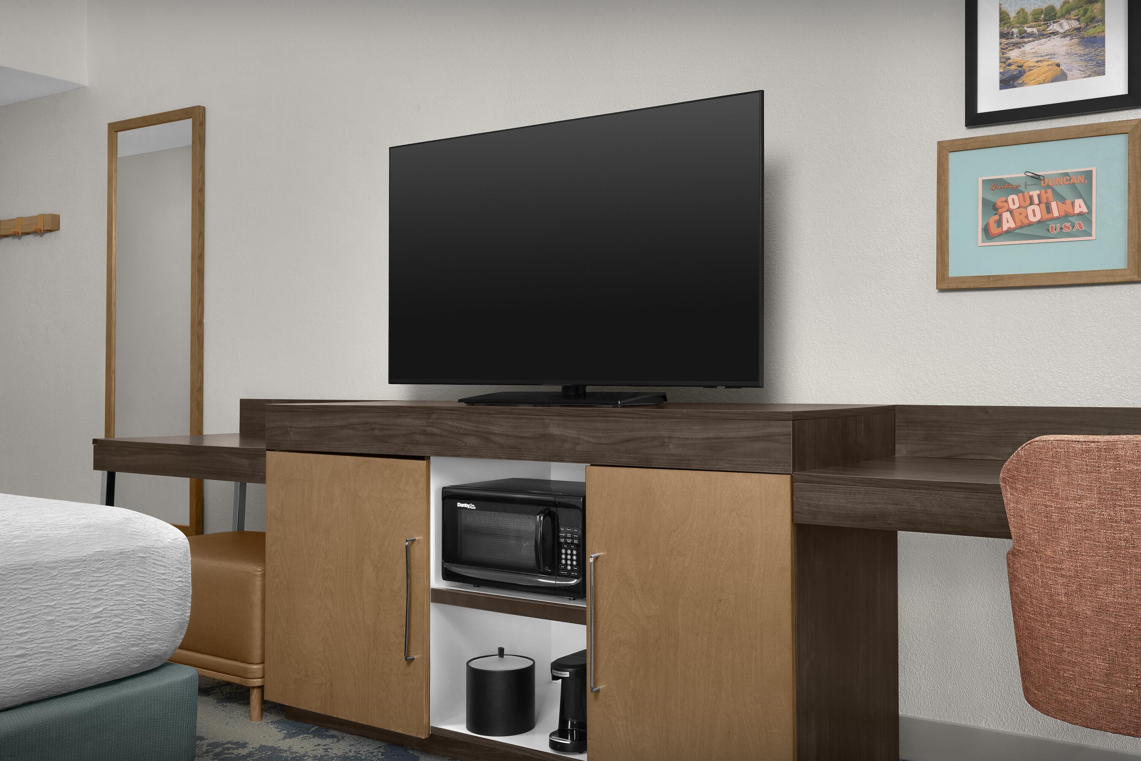 Quarto de hotel com cama, TV sobre suporte de madeira, micro-ondas e cafeteira em armário.
