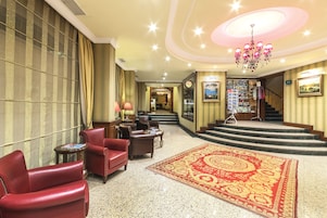 Um saguão de hotel com poltronas de couro, um carpete estampado, um candelabro e uma escada que leva aos andares superiores.