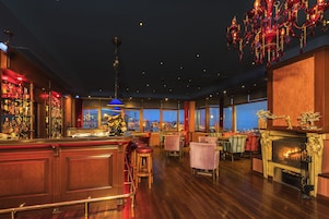 Uma área de bar com piso de madeira, balcão, banquetas e uma grande janela com vista para a paisagem urbana.