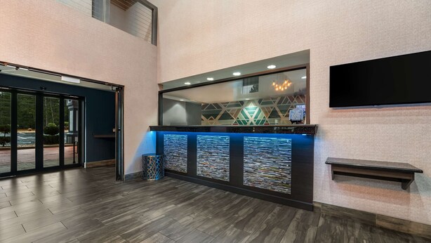 Uma área de recepção com design moderno, com um balcão de vidro e painéis iluminados por trás, uma TV de tela plana na parede e piso de madeira.