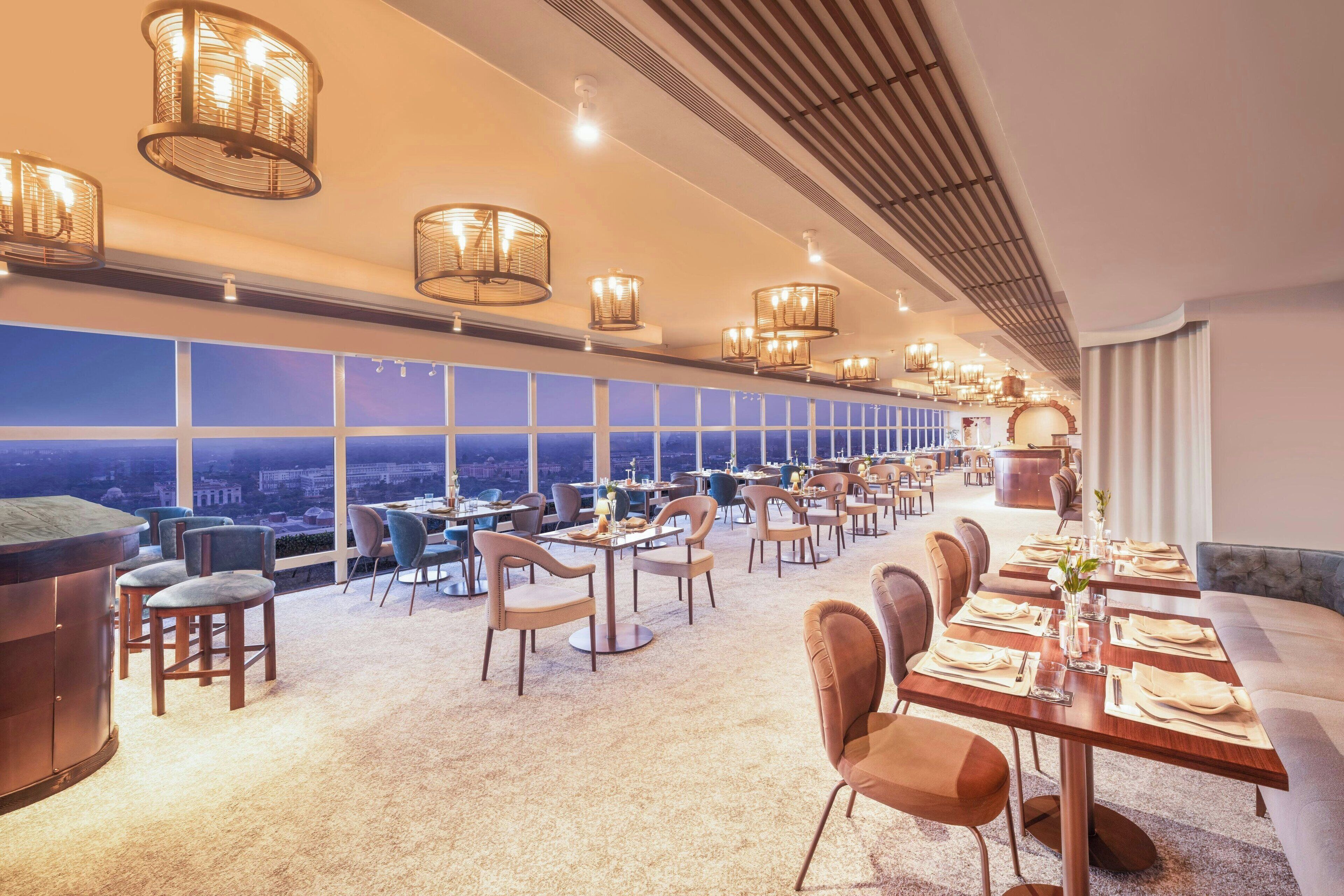 Uma sala de jantar ampla, com várias mesas e cadeiras, grandes janelas com vista para a cidade e luminárias pendentes modernas.