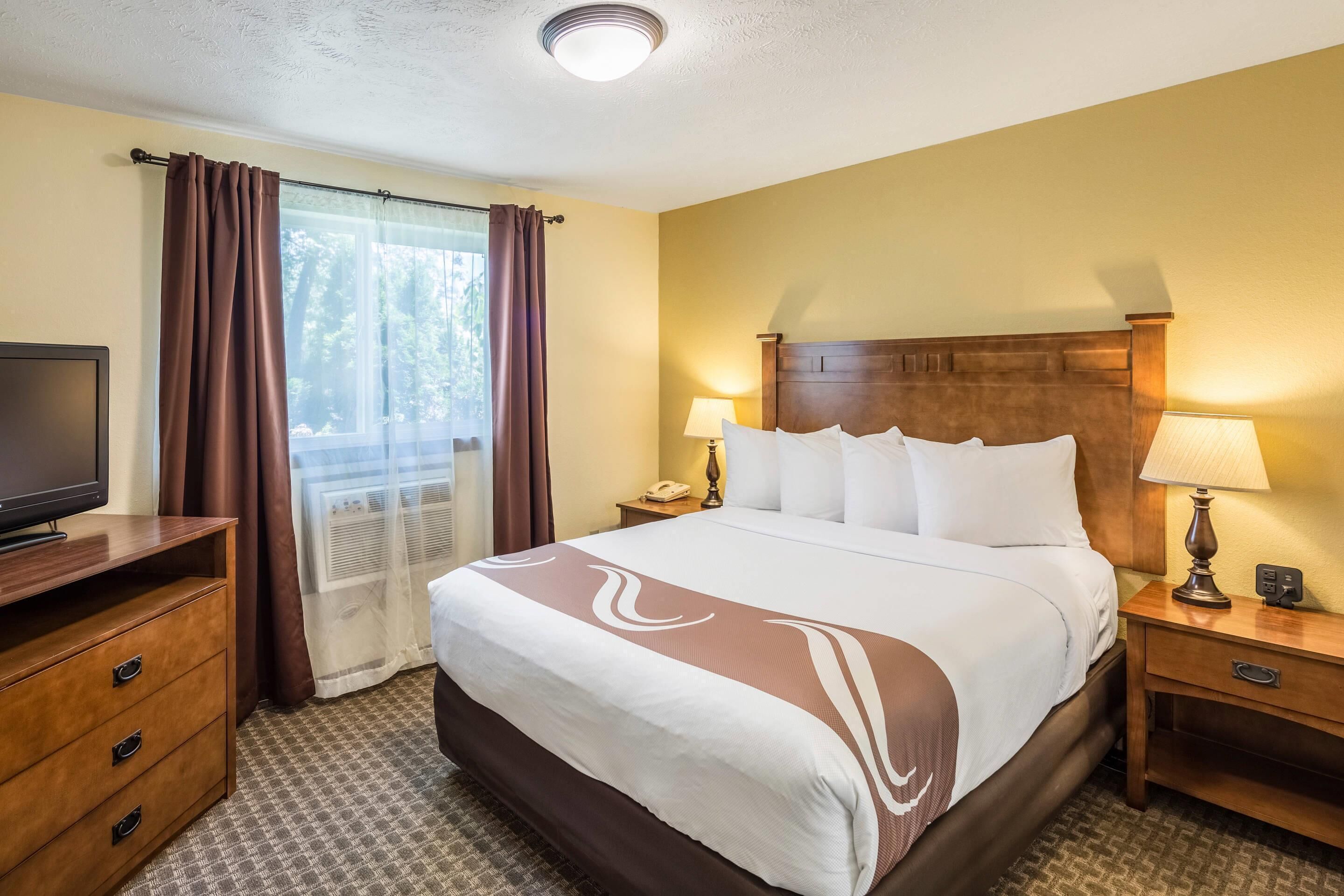 Quarto de hotel com cama, televisão, secretária, candeeiro e janela com cortinas.