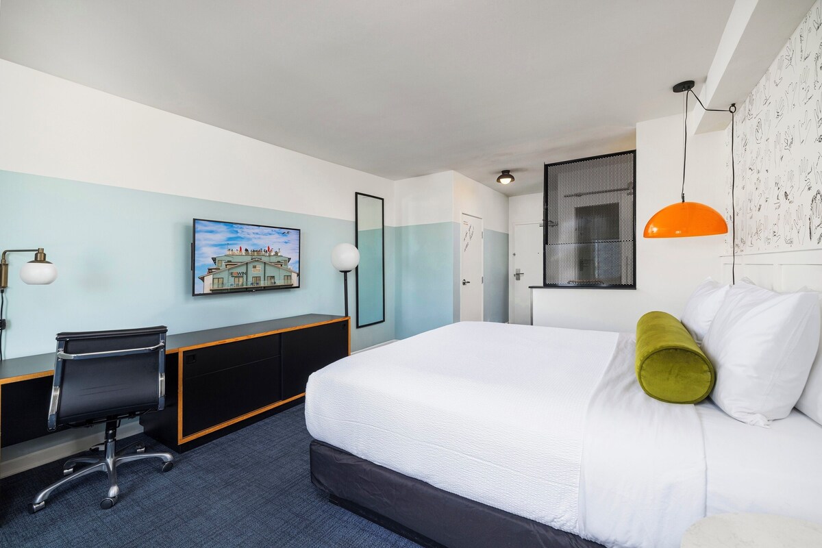 Quarto de hotel com cama, escrivaninha, cadeira e um quadro na parede.