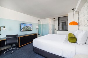 Quarto de hotel com cama, escrivaninha, cadeira e um quadro na parede.