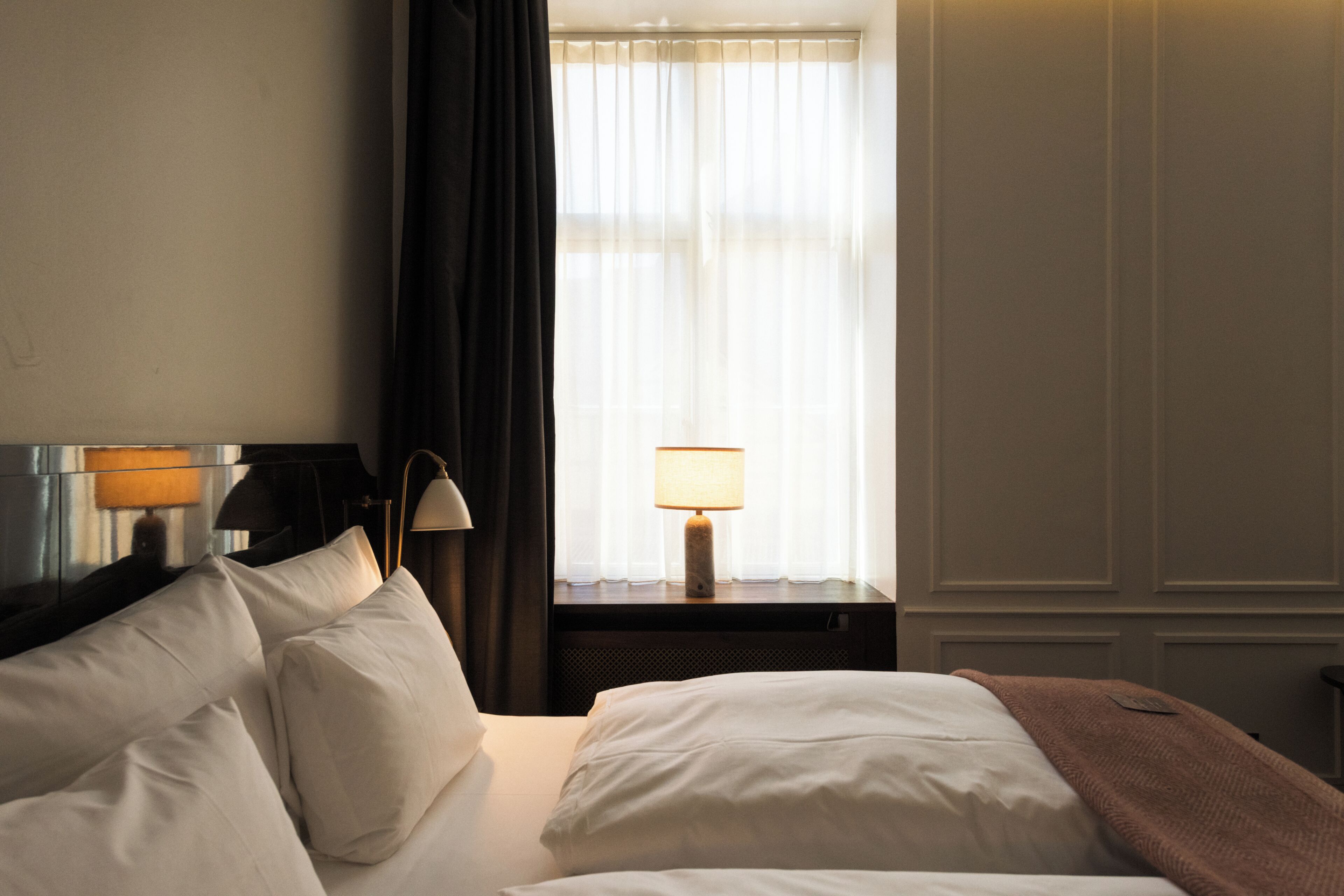 Quarto de hotel com cama, duas luminárias de cabeceira, janela com cortinas e mesa de cabeceira.