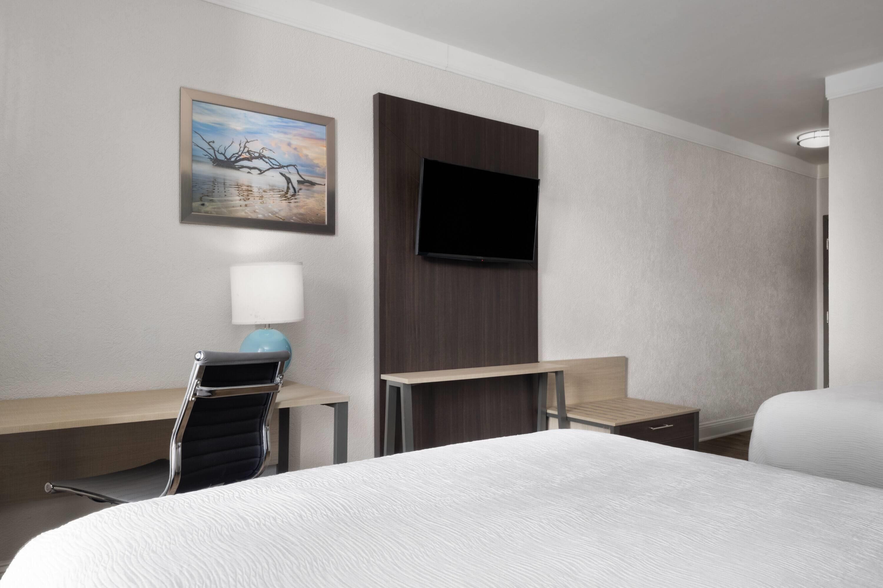 Quarto de hotel com cama, escrivaninha, cadeira, televisão na parede e um quadro.