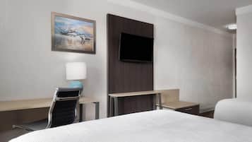 Quarto de hotel com cama, escrivaninha, cadeira, televisão na parede e um quadro.