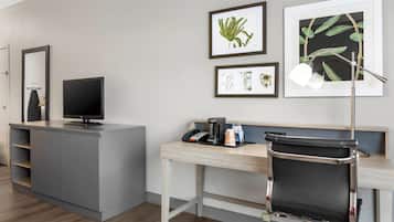 Quarto de hotel com uma escrivaninha, uma cadeira, uma televisão, uma máquina de café e obras de arte botânicas emolduradas na parede.