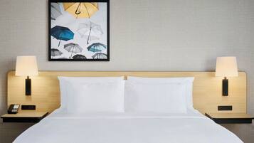 Quarto de hotel com uma cama, duas mesas de cabeceira, uma luminária na parede, um controle remoto e um quadro com uma imagem de guarda-chuvas.
